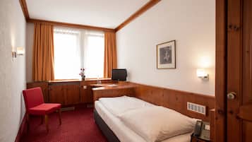 Quarto de hotel com cama, escrivaninha com computador, cadeira, telefone e janela com cortinas.