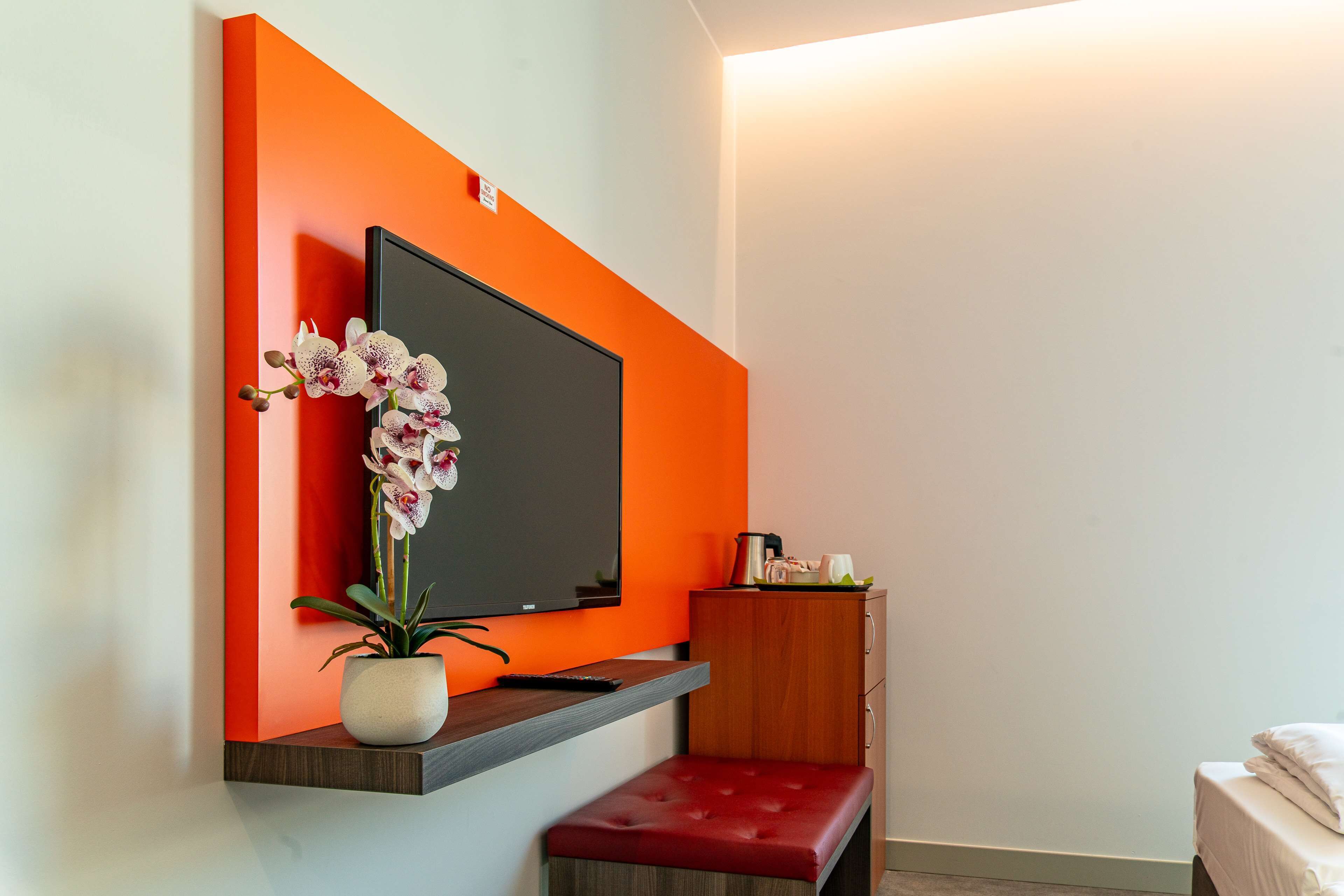 Quarto moderno com televisão presa na parede, um banco de madeira, uma pequena mesa com uma planta e uma cama com roupa de cama branca.