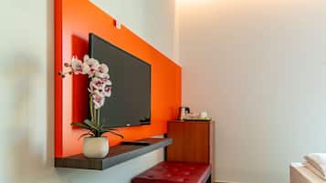 Quarto moderno com televisão presa na parede, um banco de madeira, uma pequena mesa com uma planta e uma cama com roupa de cama branca.