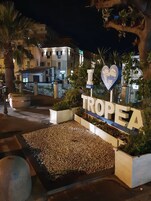 Vista noturna de uma placa com a palavra 'TROPEA' e um símbolo de coração, cercada por jardineiras e uma área de cascalho.