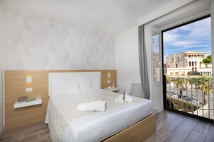 Quarto de hotel com uma cama grande, uma mesa de cabeceira, prateleira na parede e vista da rua através de uma varanda.