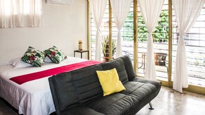 Quarto com cama, sofá cinza com travesseiro amarelo, manta vermelha e janela com cortinas.