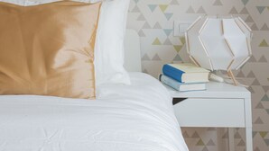 Uma cama bem arrumada com um cobertor branco e cinza listrado, um travesseiro dourado e um travesseiro branco. Uma mesa de cabeceira branca com uma decoração geométrica na parede e um abajur branco fica ao lado da cama.