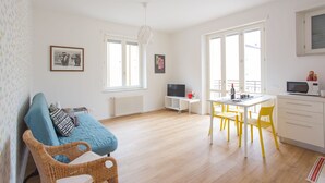 Uma sala de estar iluminada, com um sofá azul, uma cadeira de vime, uma mesa de jantar com cadeiras amarelas, uma TV e uma área de cozinha com micro-ondas.