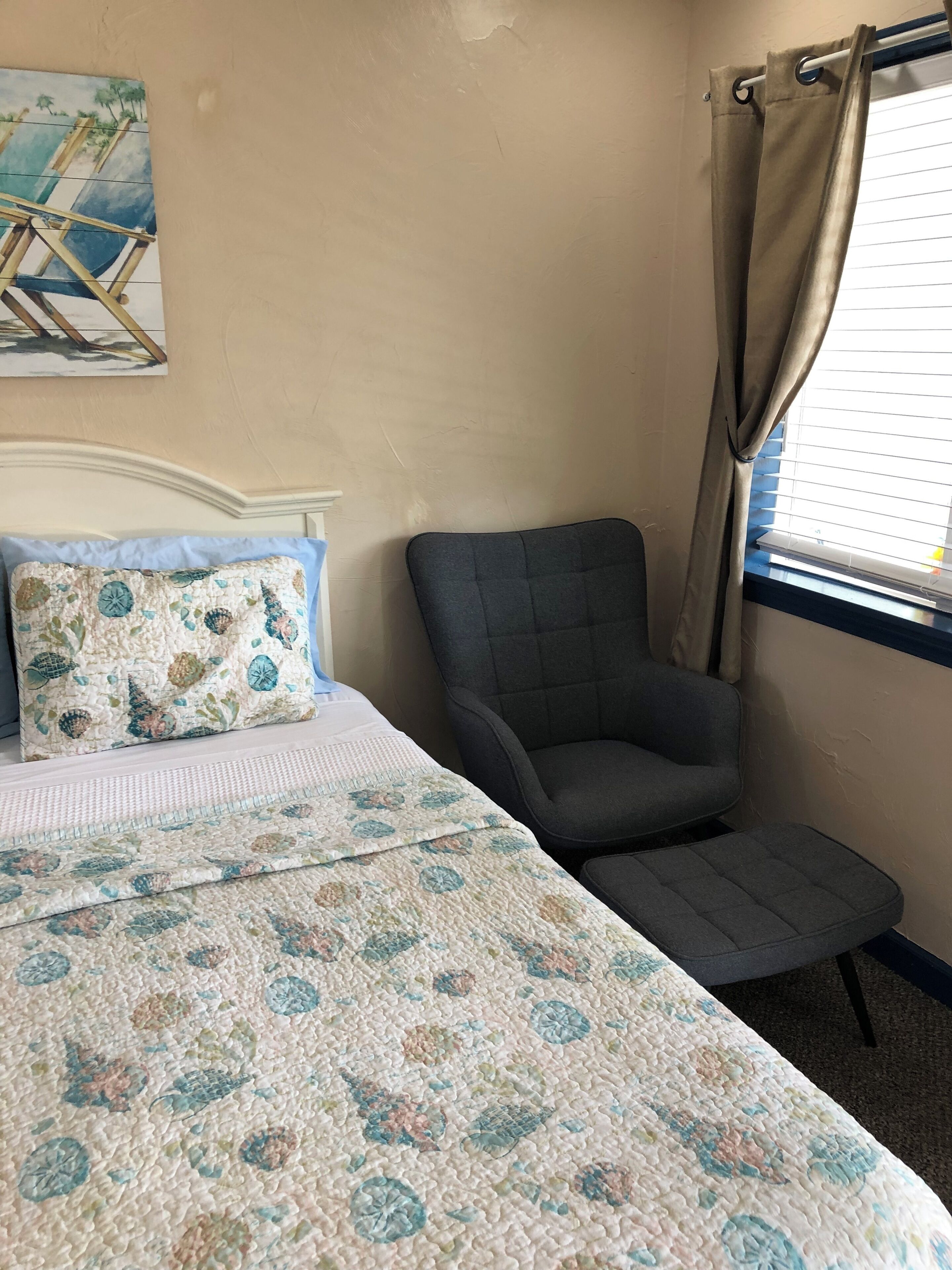 Quarto com cama, cadeira, janela com cortinas e um quadro na parede.