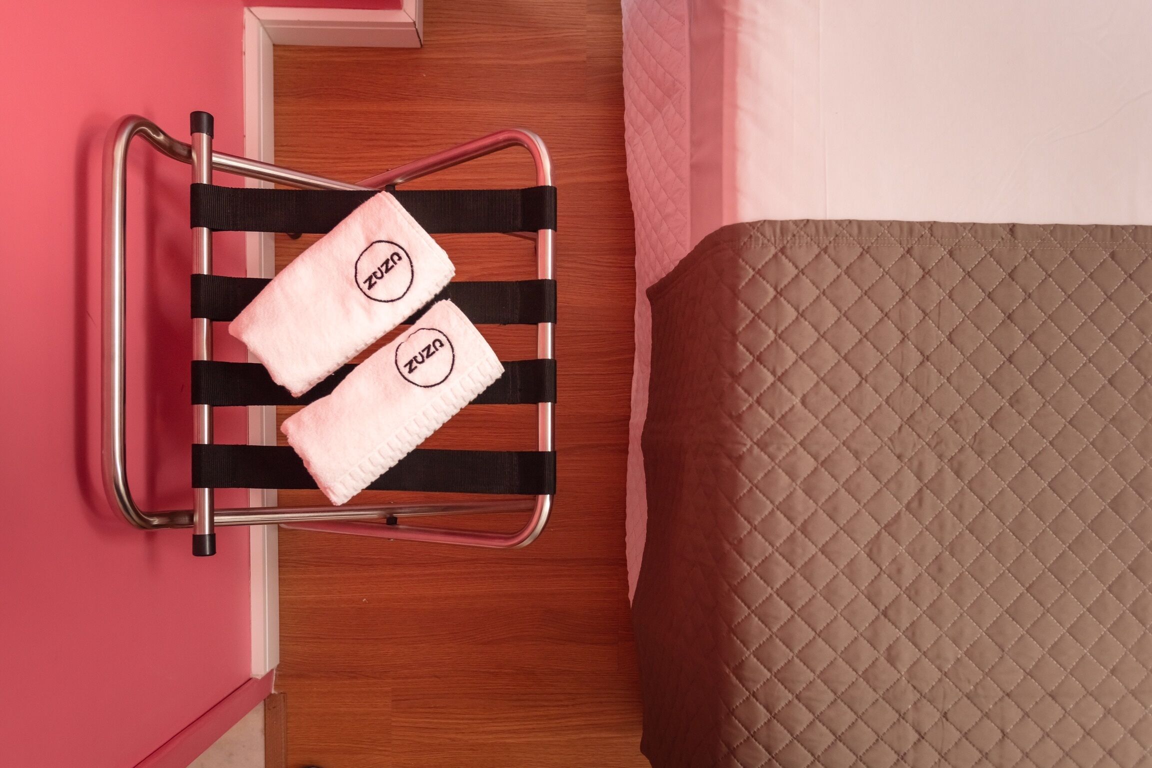Uma cama com cobertura acolchoada, um suporte de metal com duas toalhas e uma parede rosa.