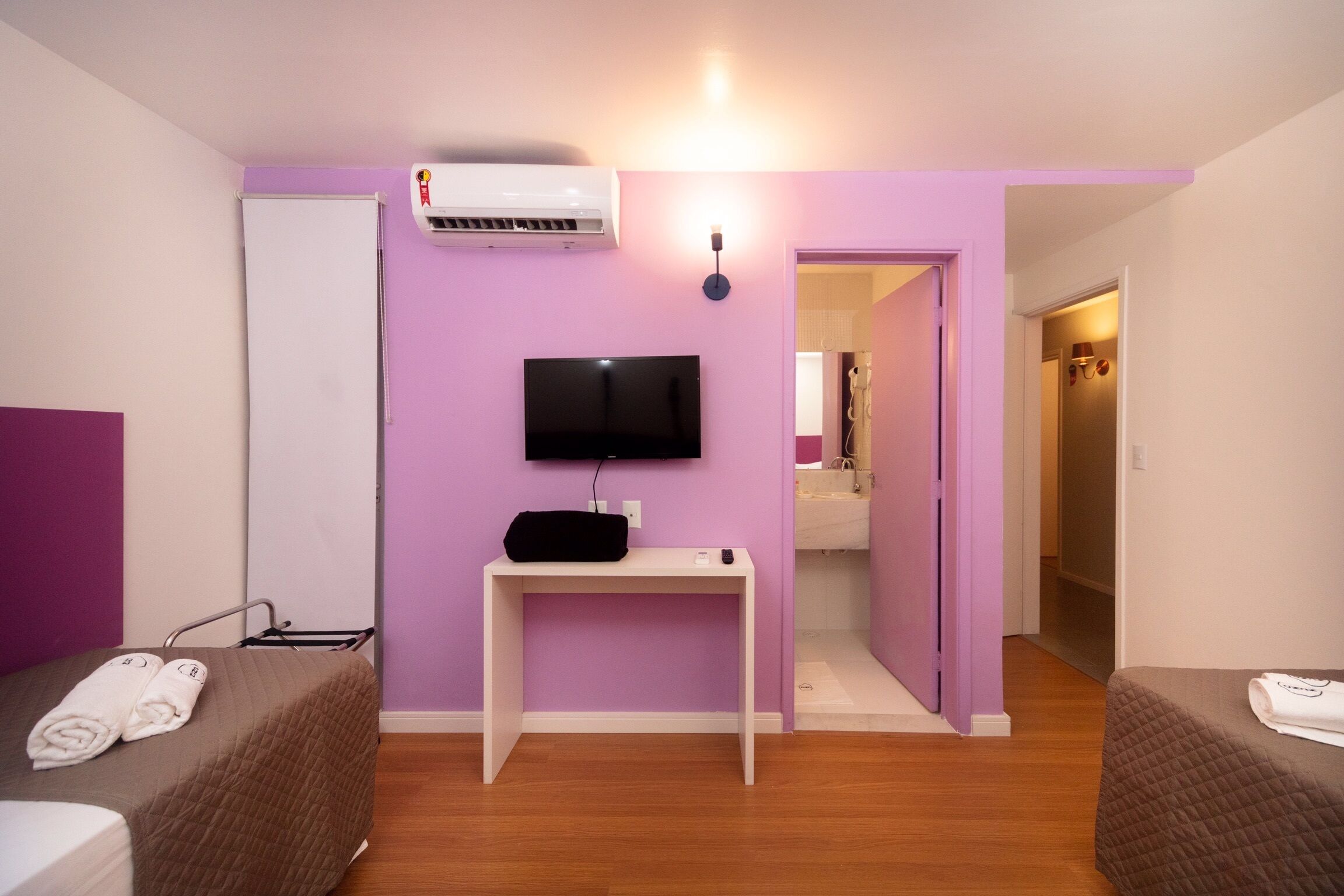Quarto com uma cama, uma mesinha com televisão, ar condicionado e um banheiro visível por uma porta aberta.