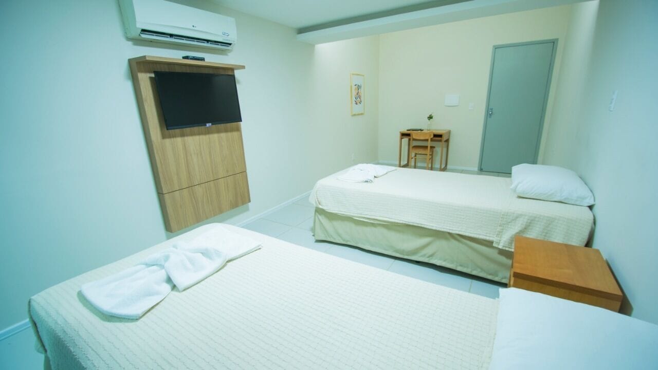 Quarto de hotel com duas camas, uma televisão presa a um móvel de madeira, uma escrivaninha com uma cadeira e uma porta.