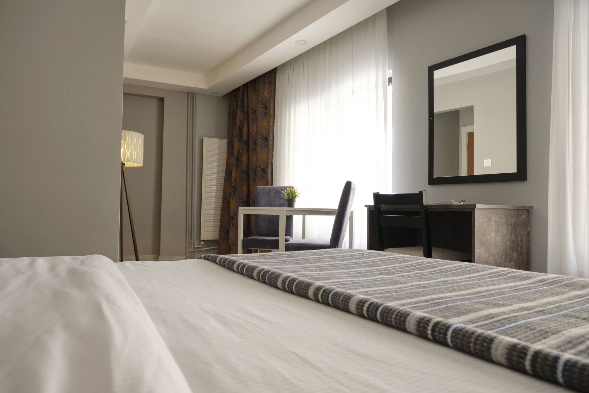 Quarto de hotel com uma cama grande, uma escrivaninha com cadeira, uma mesinha, um espelho e um abajur.