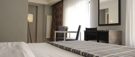 Quarto de hotel com uma cama grande, uma escrivaninha com cadeira, uma mesinha, um espelho e um abajur.