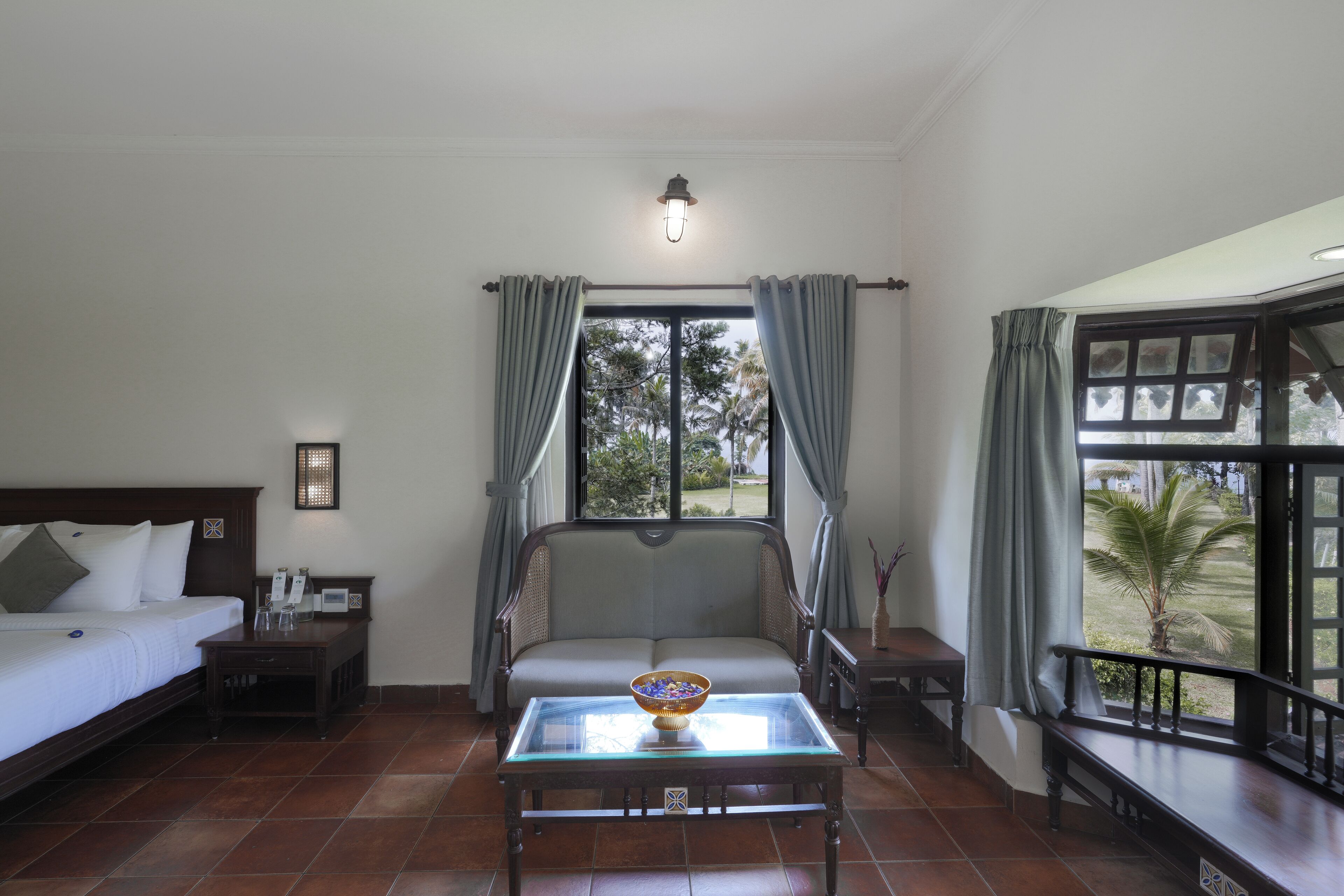 Quarto de hotel com cama, sofá, mesa de centro de vidro com uma tigela decorativa, mesa de cabeceira, janela com cortinas e vista para palmeiras.