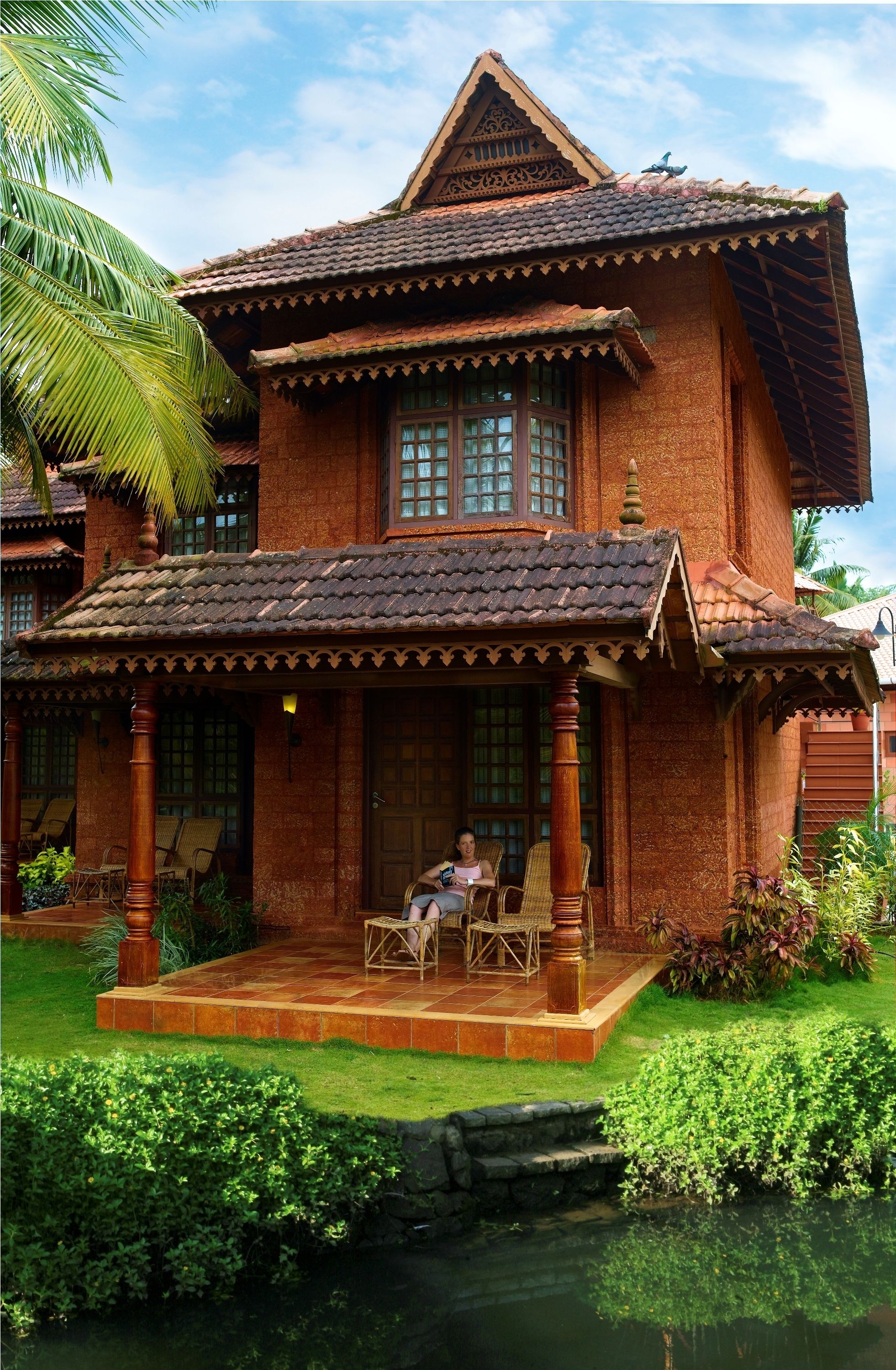 Uma casa tradicional com telhado de telhas, varanda com cadeiras e um pequeno lago ao primeiro plano.