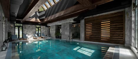 Piscina interior moderna com claraboia, paredes de pedra e vigas de madeira.