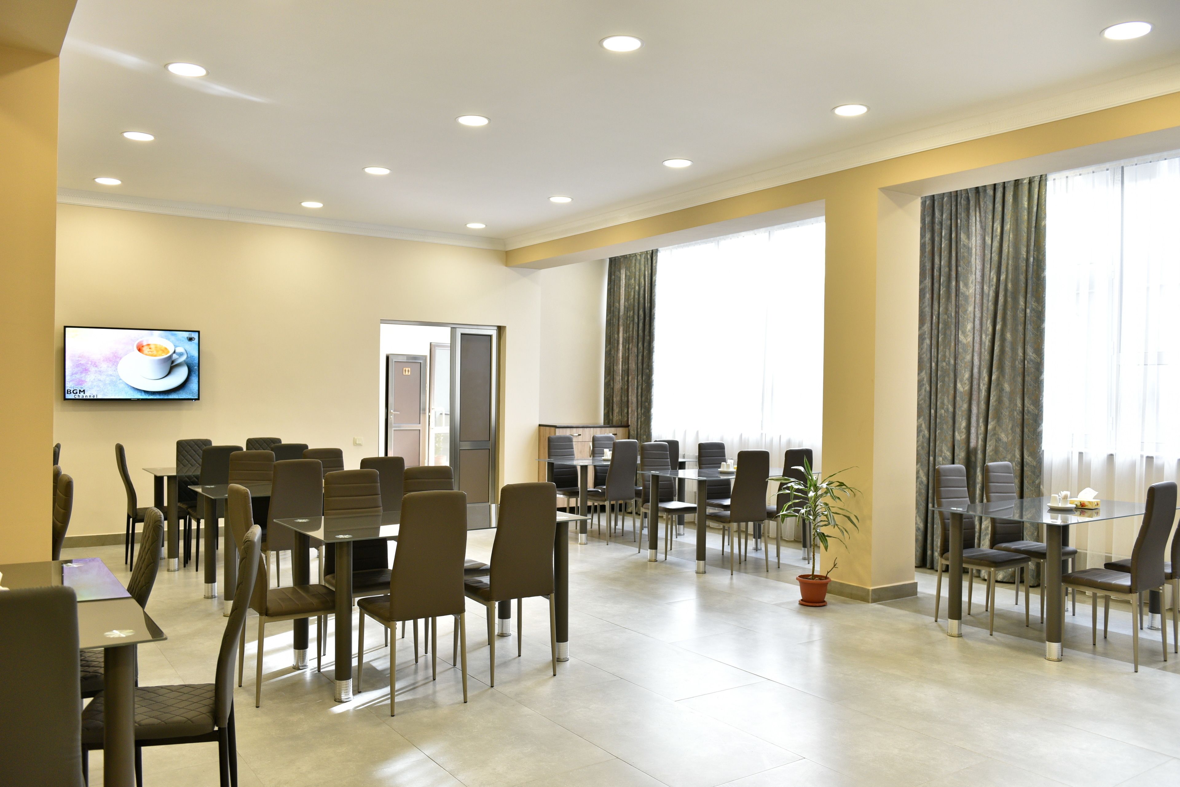 Sala de conferências com várias mesas e cadeiras, uma tela de televisão e janelas amplas com cortinas.