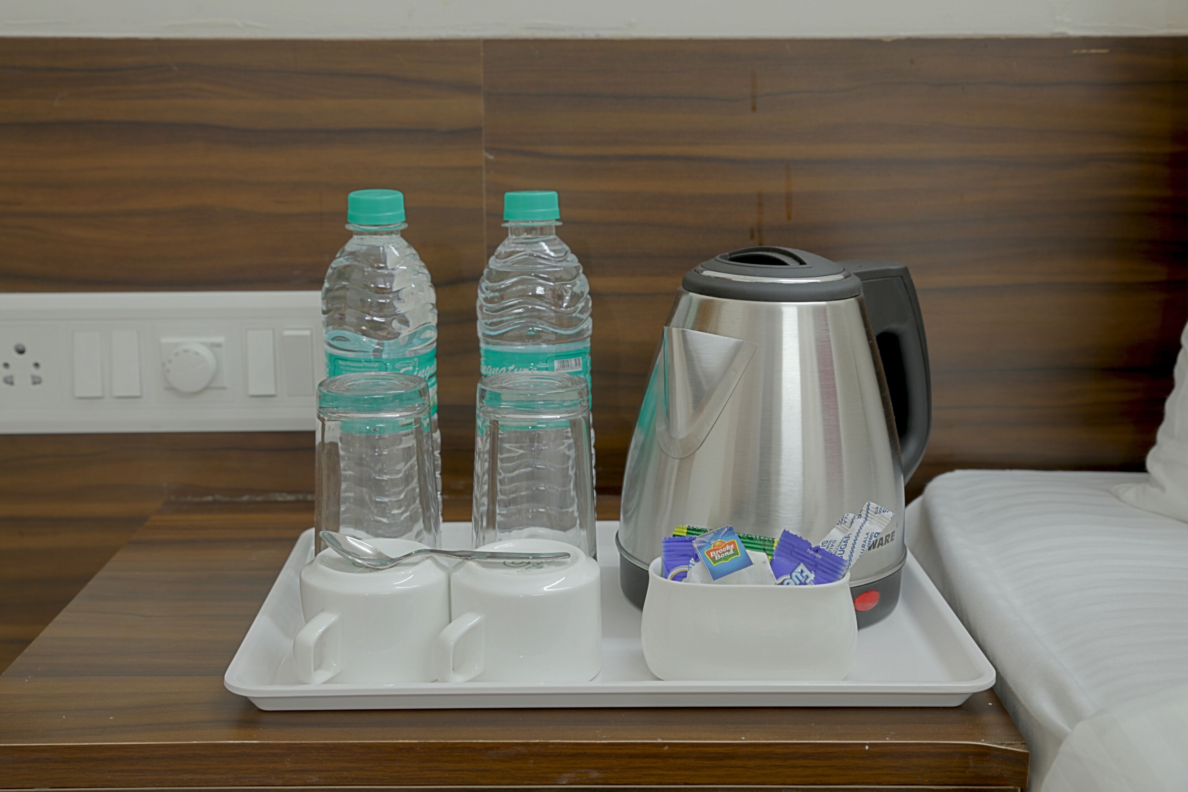 Uma bandeja com duas garrafas de água, uma cafeteira e outra bandeja com sachês sobre uma mesa de madeira em um quarto de hotel.