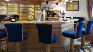 Uma área de bar com banquetas de madeira, um balcão com um jarro branco e prateleiras com copos.