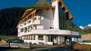 Um hotel de dois andares com montanhas ao fundo, com varandas, janelas amplas e uma placa com a inscrição 'CASADA HOTEL'.