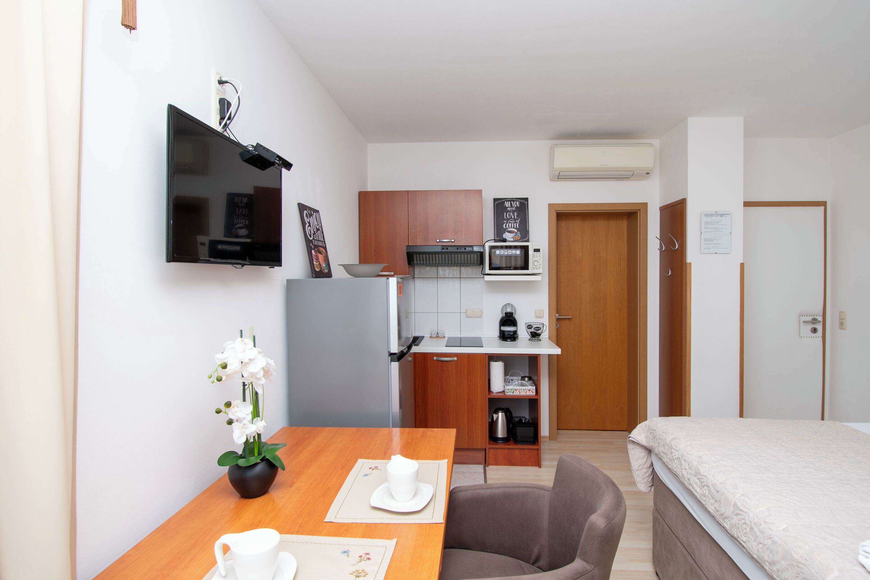 Quarto de hotel compacto com cozinha compacta, área de refeições com mesa e cadeiras, cama e um pequeno vaso de planta.