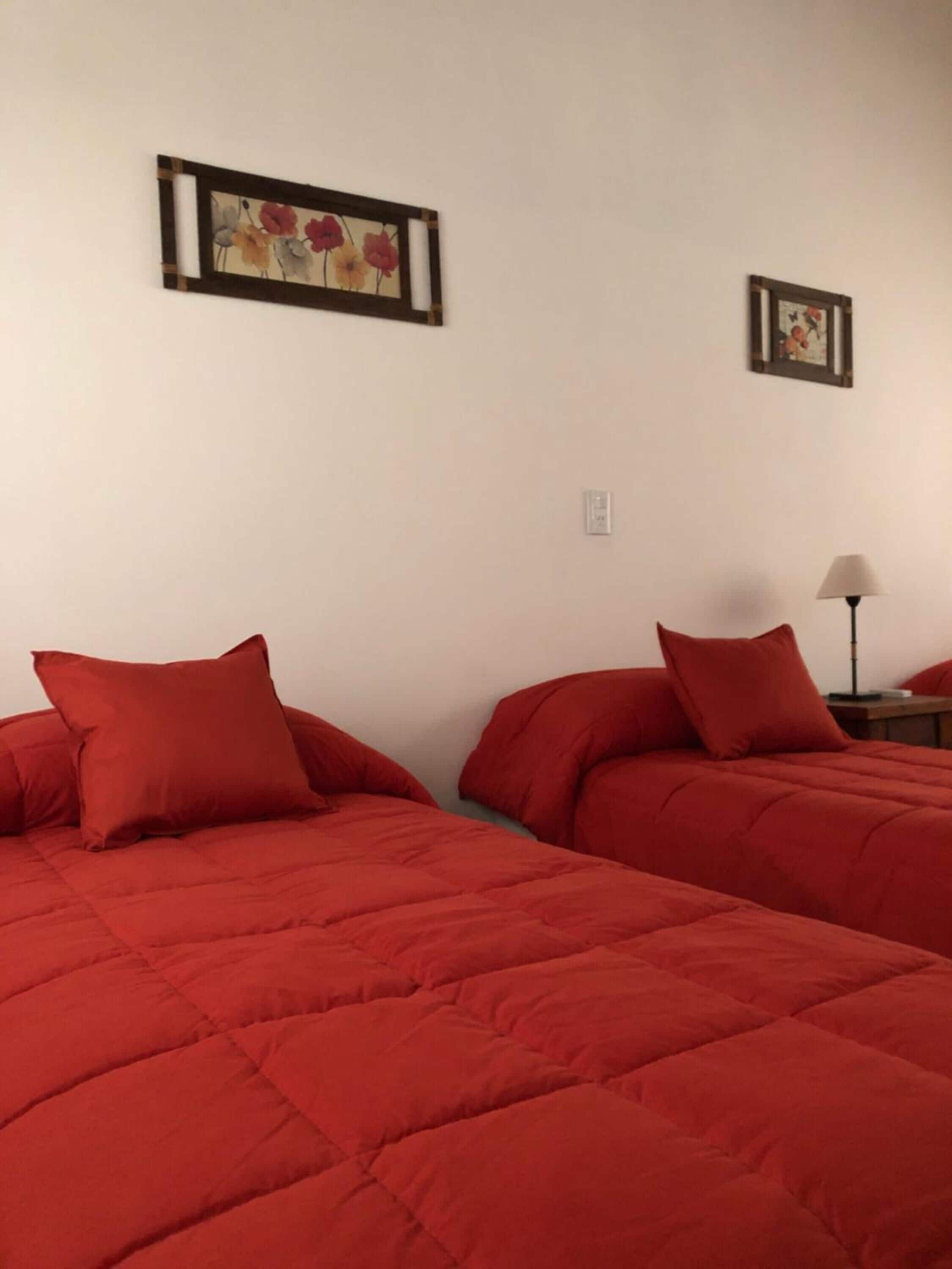 Quarto com duas camas, cada uma com roupa de cama e almofadas vermelhas. Existem duas imagens emolduradas na parede, uma acima da cama à esquerda e outra acima da cama à direita. Uma luminária é visível em uma mesa de cabeceira ao lado da cama direita.