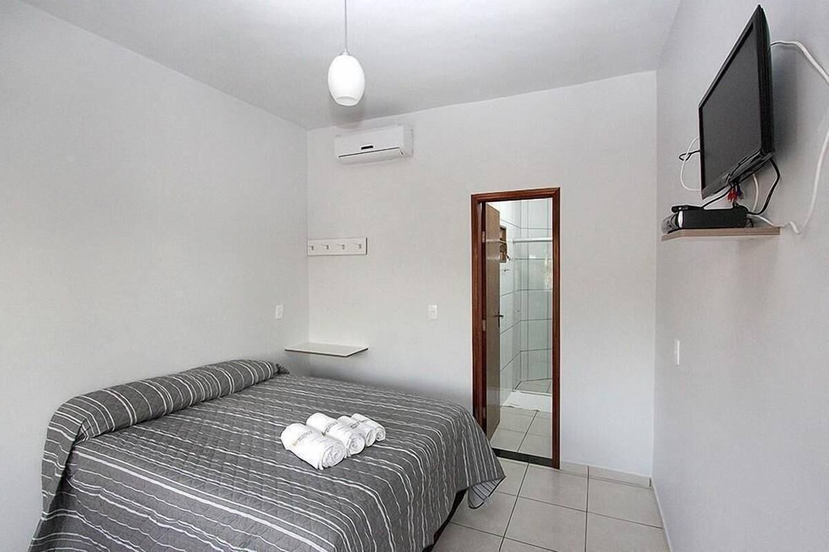 Quarto com cama, televisão sobre suporte e porta para um banheiro.