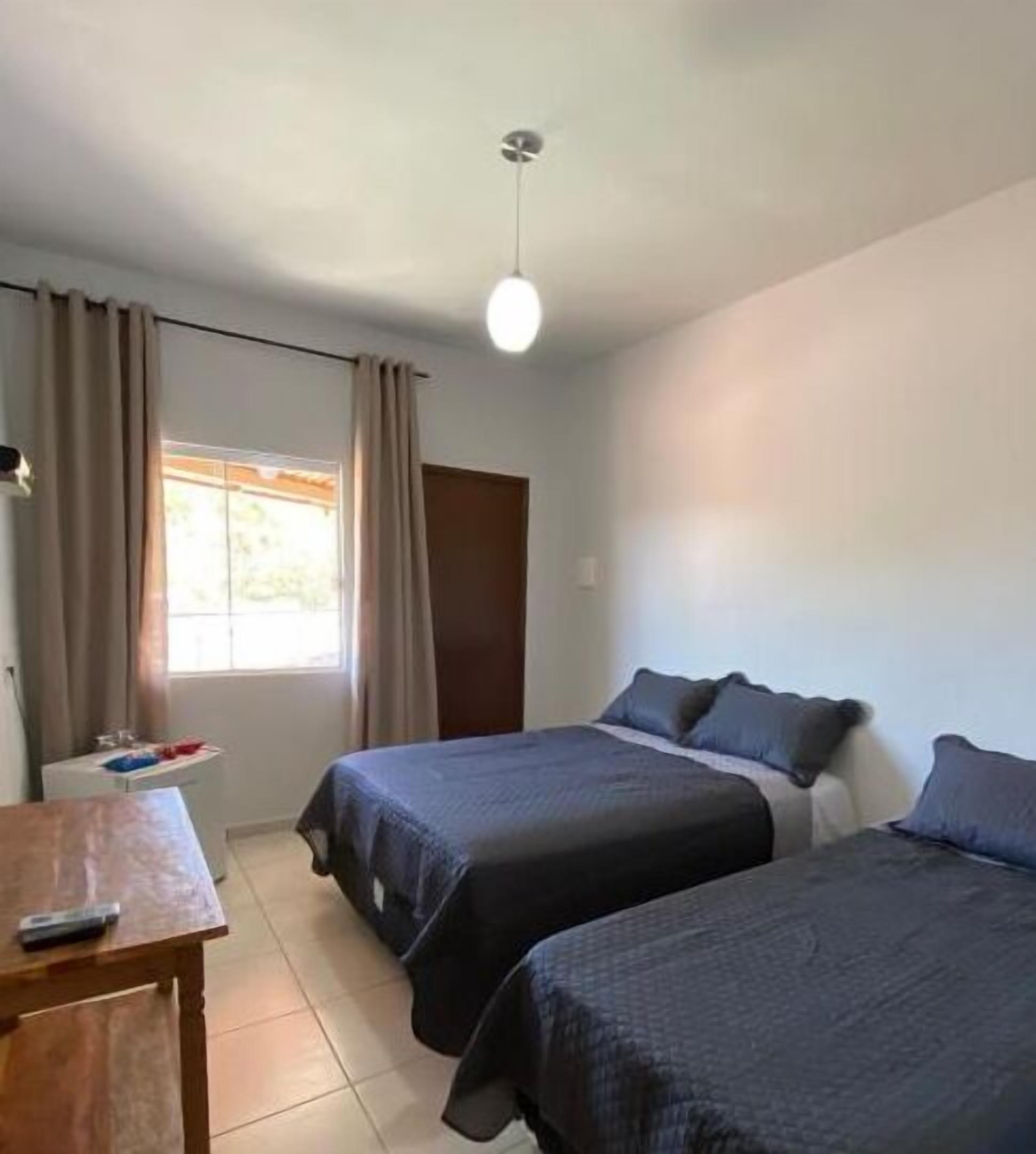 Quarto com duas camas, uma mesa pequena, uma janela com cortinas e uma luminária no teto.