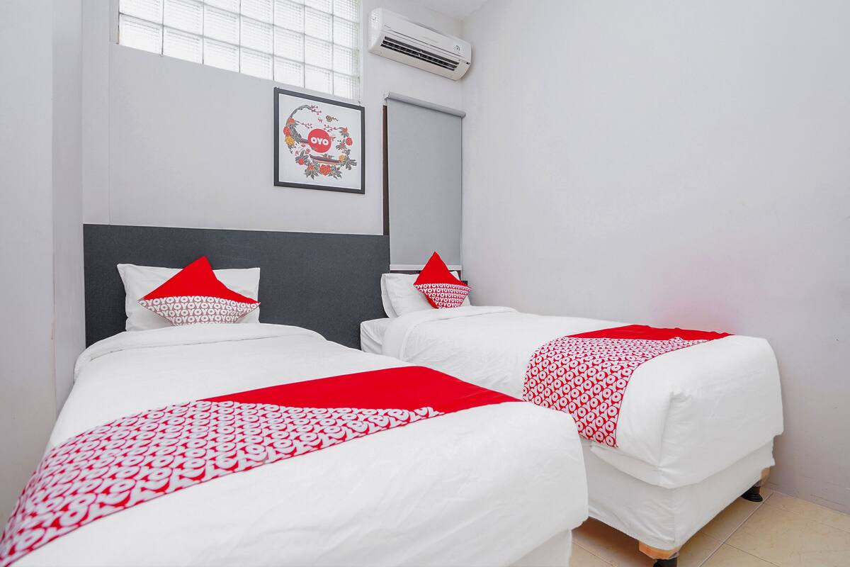 Quarto de hotel com duas camas, cada uma com roupa de cama branca e detalhes vermelhos, uma pintura floral emoldurada na parede e um aparelho de ar condicionado sobre as camas.