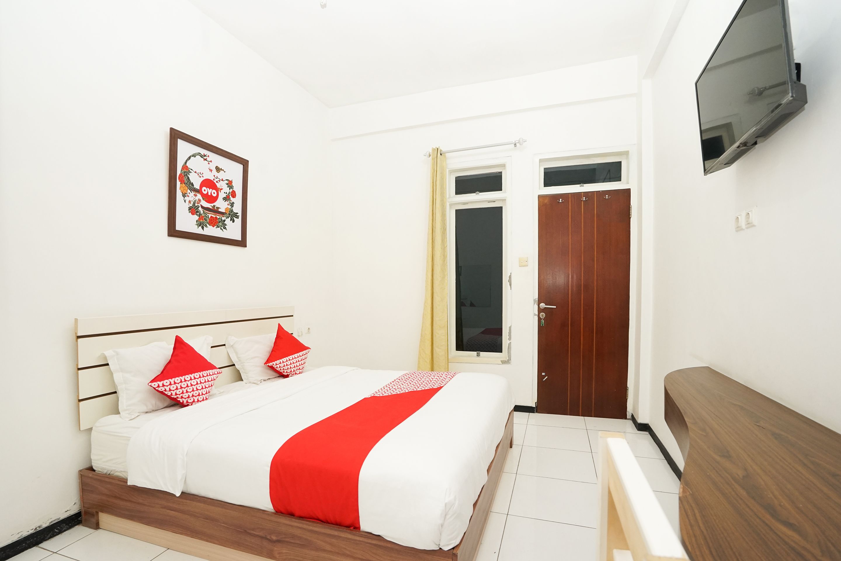 Quarto bem arrumado com cama, um banco de madeira, TV fixada na parede e porta que dá para outro cômodo.