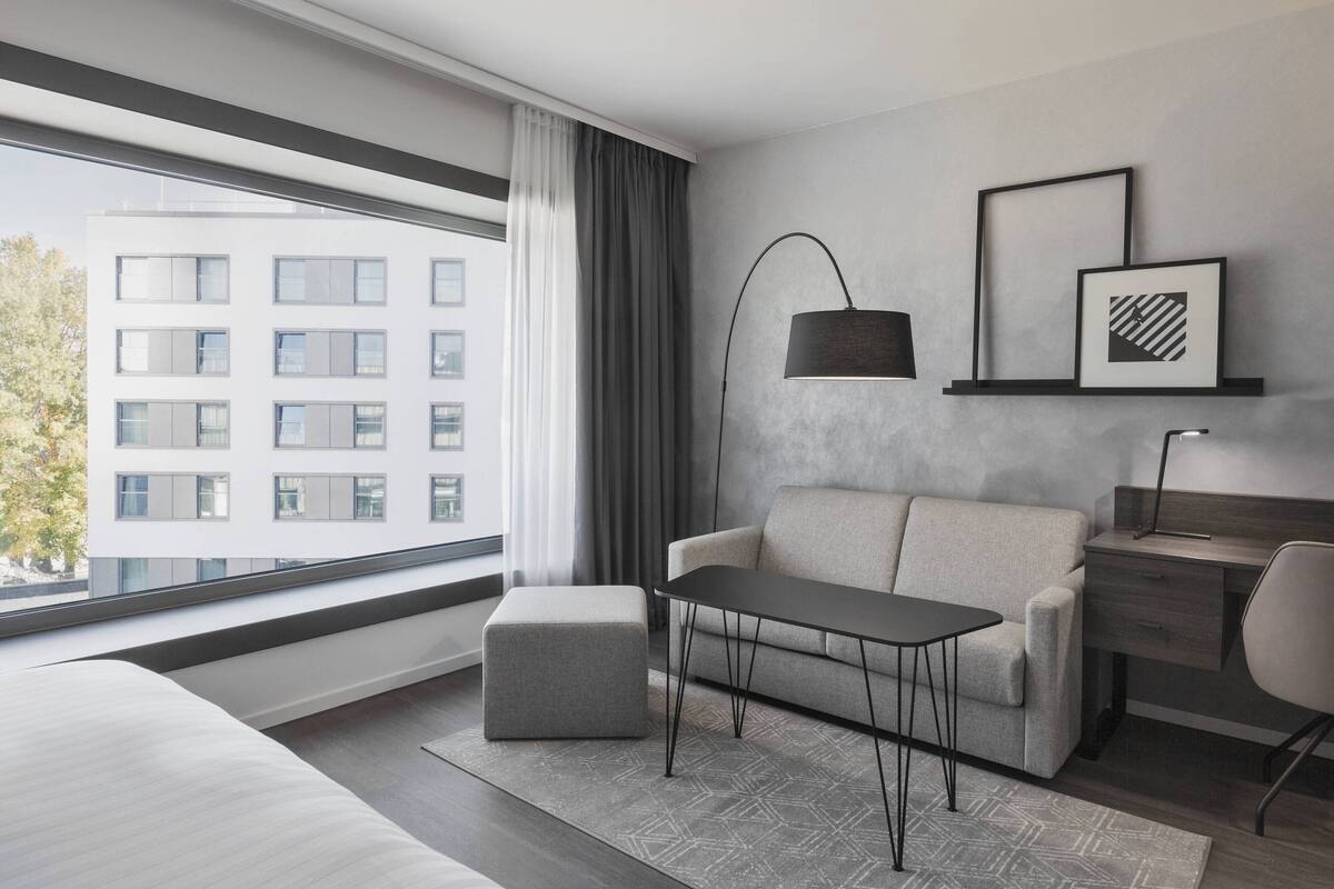 Quarto de hotel moderno com uma janela grande, um sofá, uma mesa pequena, uma escrivaninha e uma cadeira.