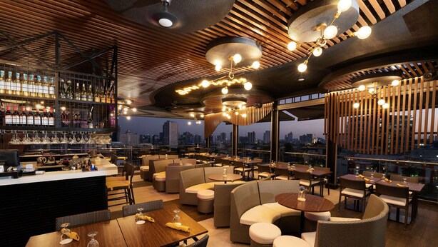Uma área de bar moderna com vista para a cidade, apresentando painéis de teto de madeira, mesas redondas e banquetas de bar.