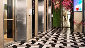 Um corredor moderno com chão xadrez preto e branco, elevadores e uma parede verde com flores cor-de-rosa.