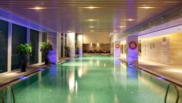 Piscina interior moderna com água cristalina, rodeada por paredes de vidro e plantas em vasos.