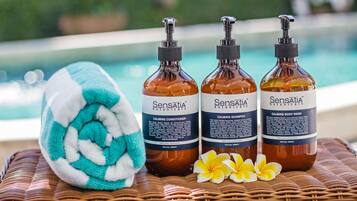 Três frascos de produtos Sensatia Botanicals, uma toalha enrolada e duas flores sobre uma superfície de vime junto a uma piscina.