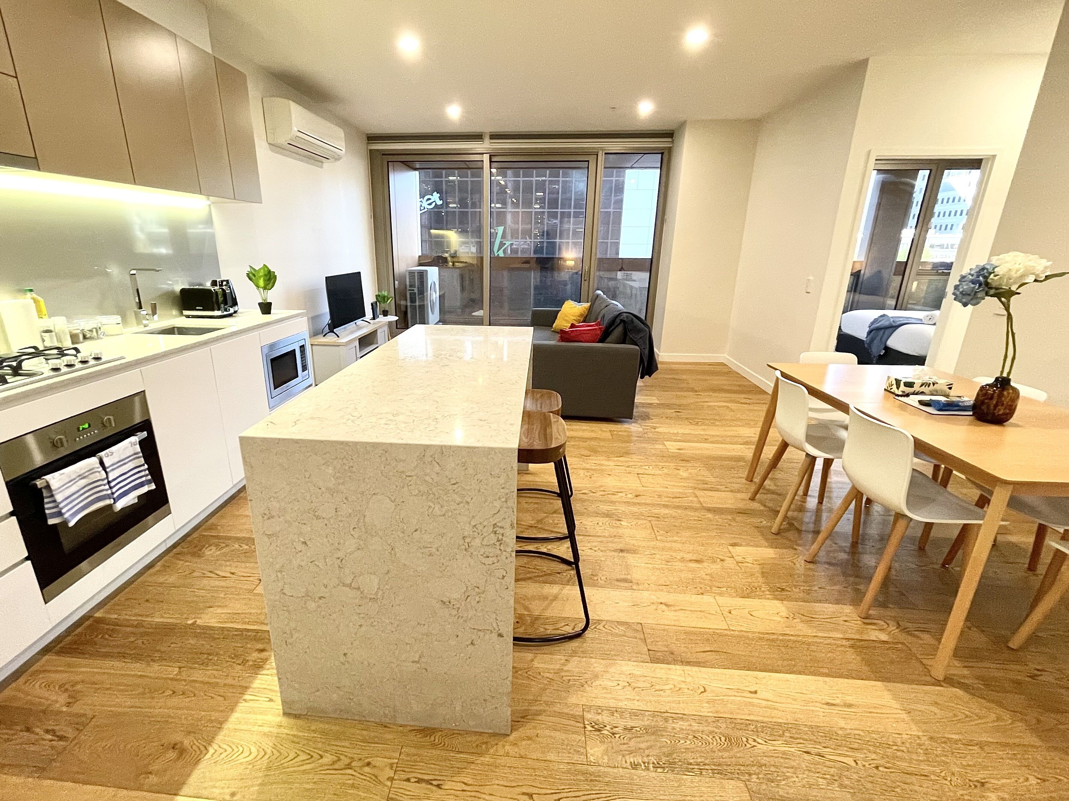 Cozinha moderna com ilha central, área de refeições com mesa e cadeiras de madeira e sala de estar com sofá e janela ampla.