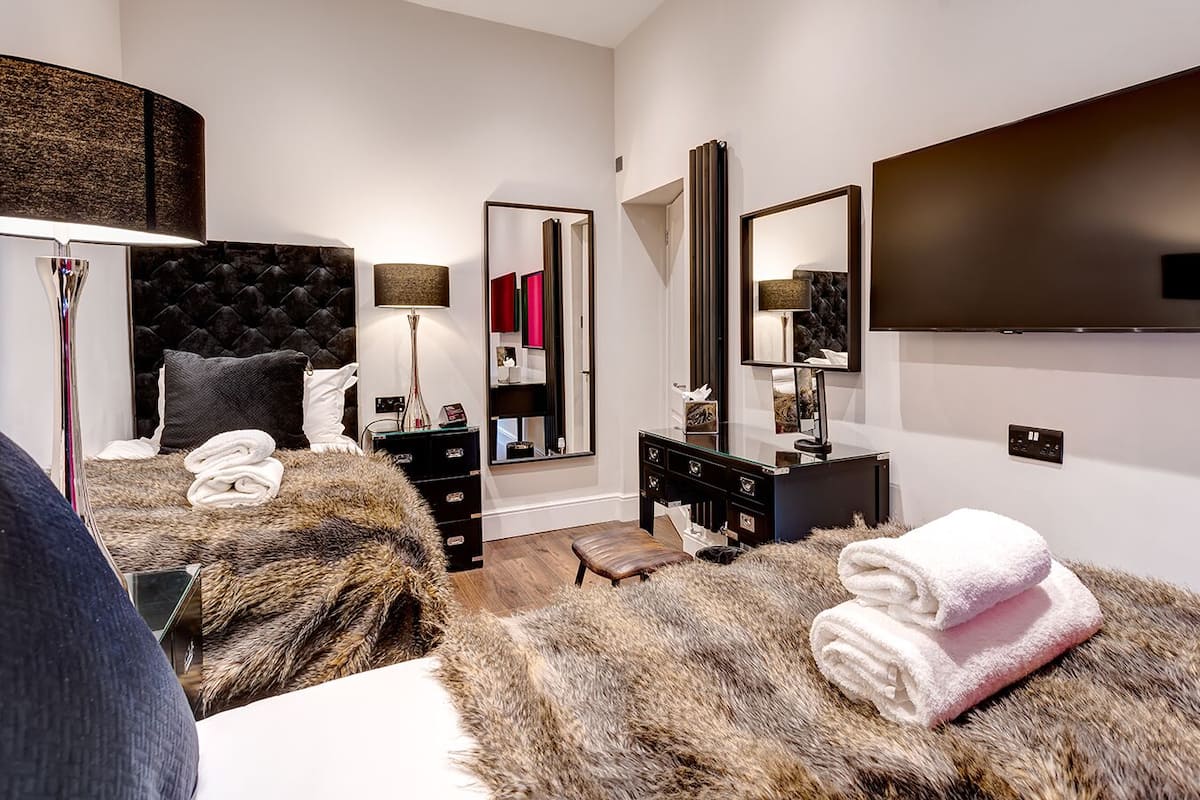 Quarto moderno com uma cama grande, uma toucador com espelho, uma TV de tela plana na parede e um banquinho revestido de pele.