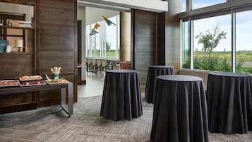Uma sala de conferência moderna com mesas redondas cobertas por toalhas pretas, uma mesa de buffet com comida e grandes janelas com vista para o exterior.