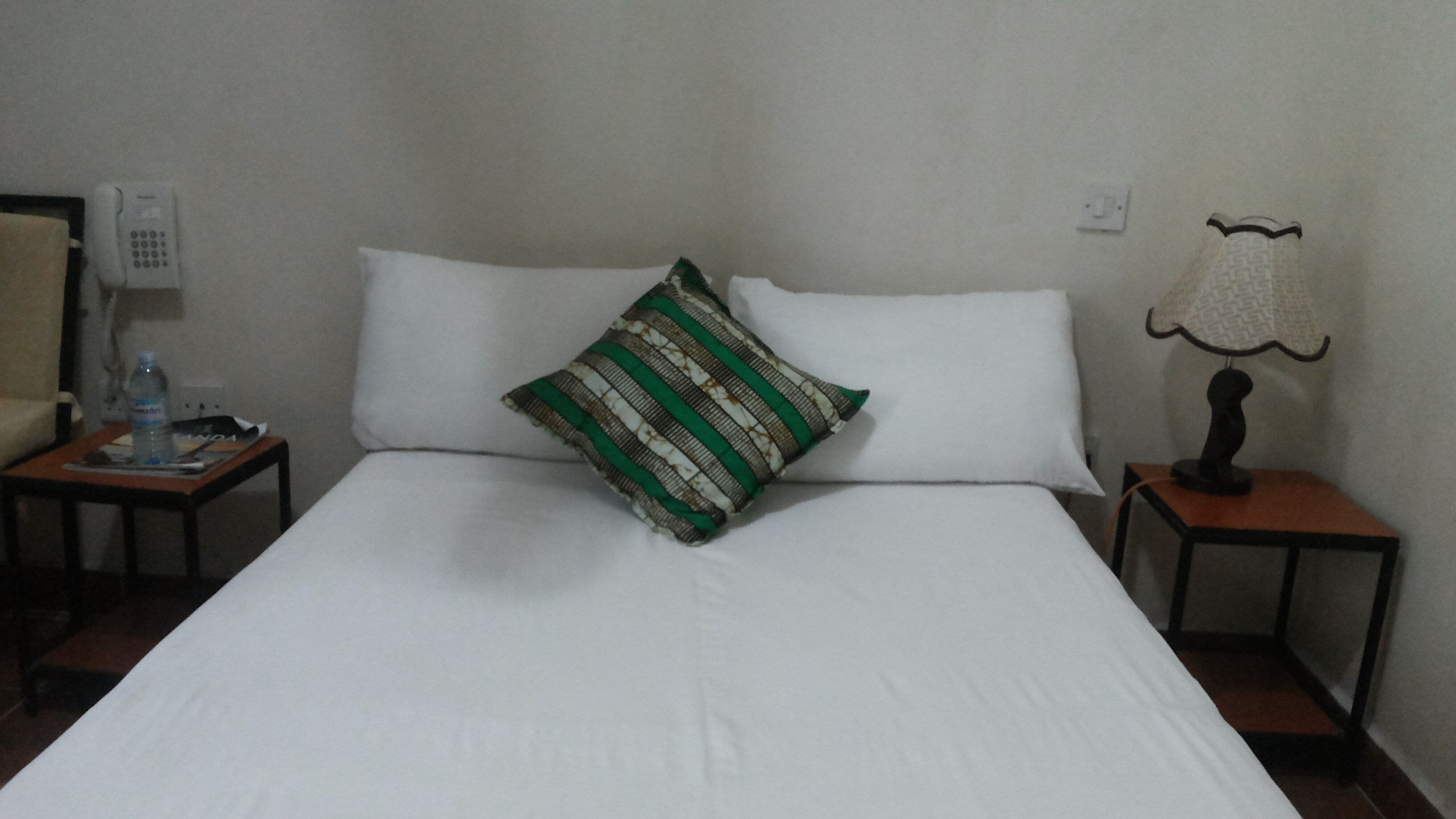 Cama bem arrumada com lençóis brancos e travesseiro listrado em verde, abajur, telefone e uma garrafa de água sobre uma pequena mesa ao lado da cama.