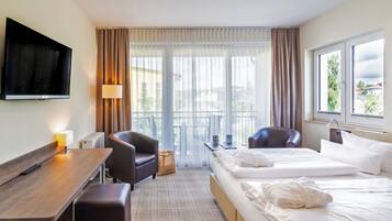 Quarto de hotel com uma cama, duas cadeiras, uma escrivaninha, uma televisão e uma janela com cortinas.