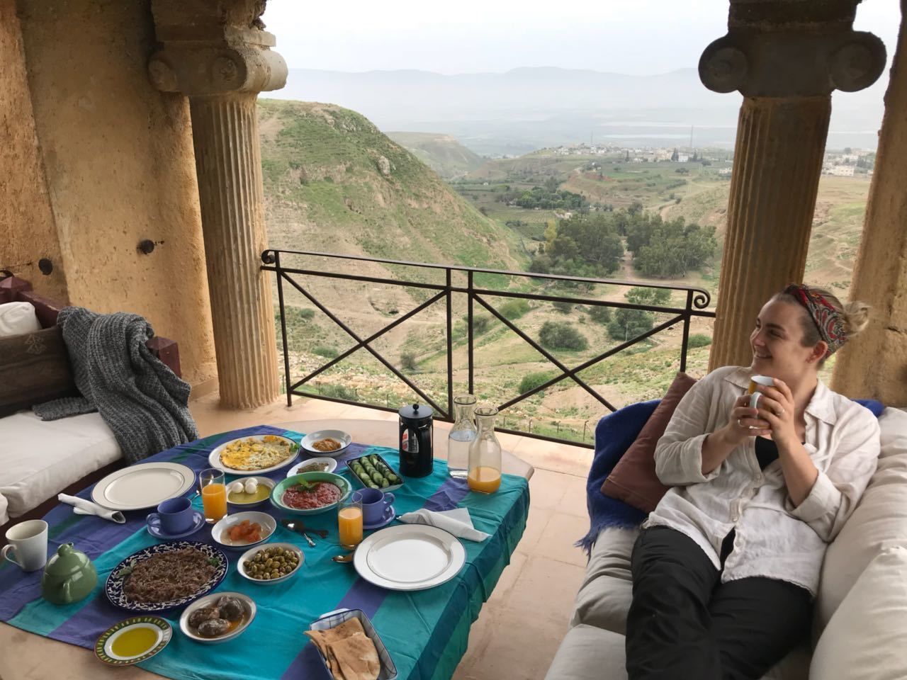 Uma mesa de jantar posta com diversos pratos e bebidas, uma pessoa sentada num sofá, e uma vista panorâmica de colinas e uma cidade ao longe.
