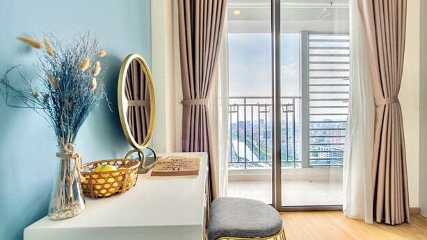 Quarto com escrivaninha branca, um vaso com plantas secas, um cesto e um espelho. Há uma janela com cortinas, uma varanda com guarda-corpo e vista para edifícios.