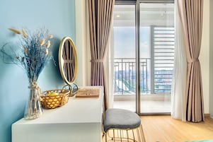 Quarto com escrivaninha branca, um vaso com plantas secas, um cesto e um espelho. Há uma janela com cortinas, uma varanda com guarda-corpo e vista para edifícios.