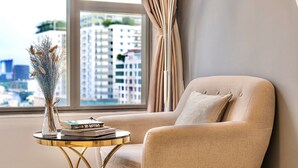 Uma sala de estar moderna com vista para o horizonte urbano, uma poltrona bege, uma pequena mesa redonda com livros e um vaso, e um banquinho amarelo.