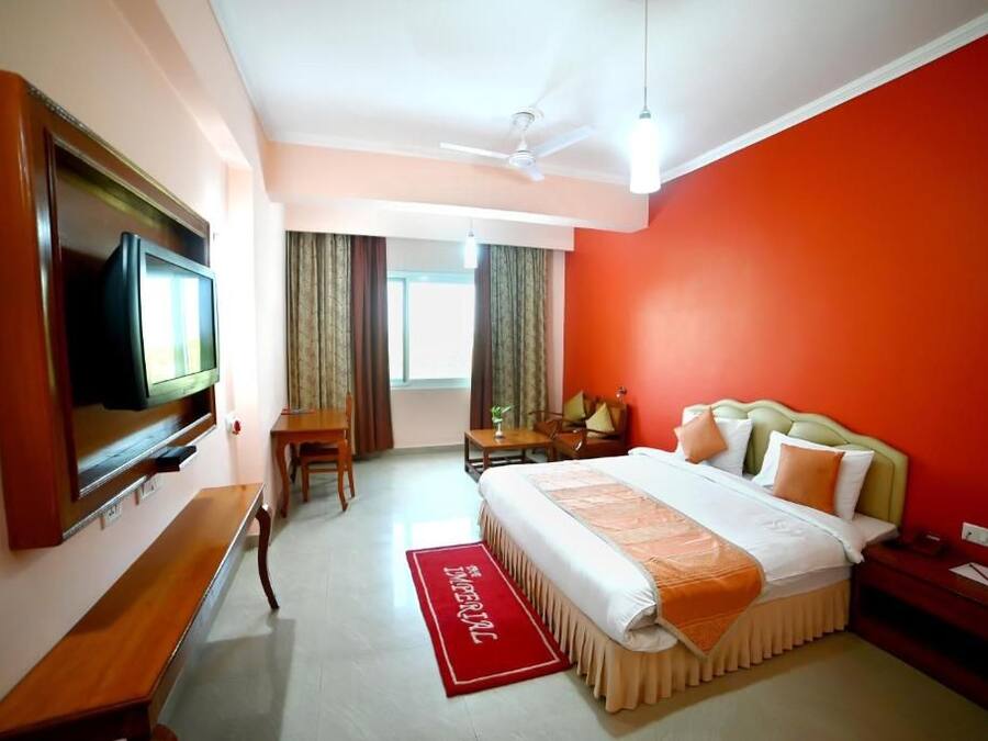 Quarto de hotel com cama, escrivaninha, cadeira, televisão e tapete vermelho.
