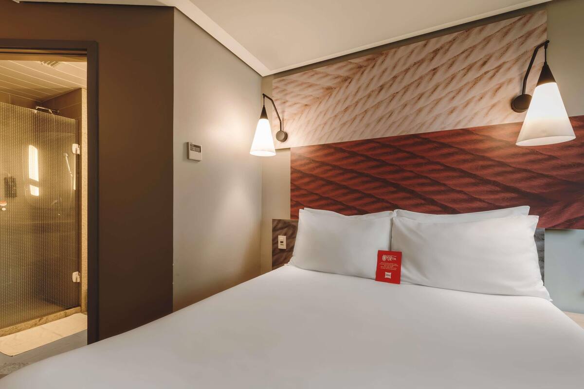 Quarto de hotel com cama, duas luminárias fixadas na parede, cabeceira de madeira e um sinal vermelho sobre os travesseiros.