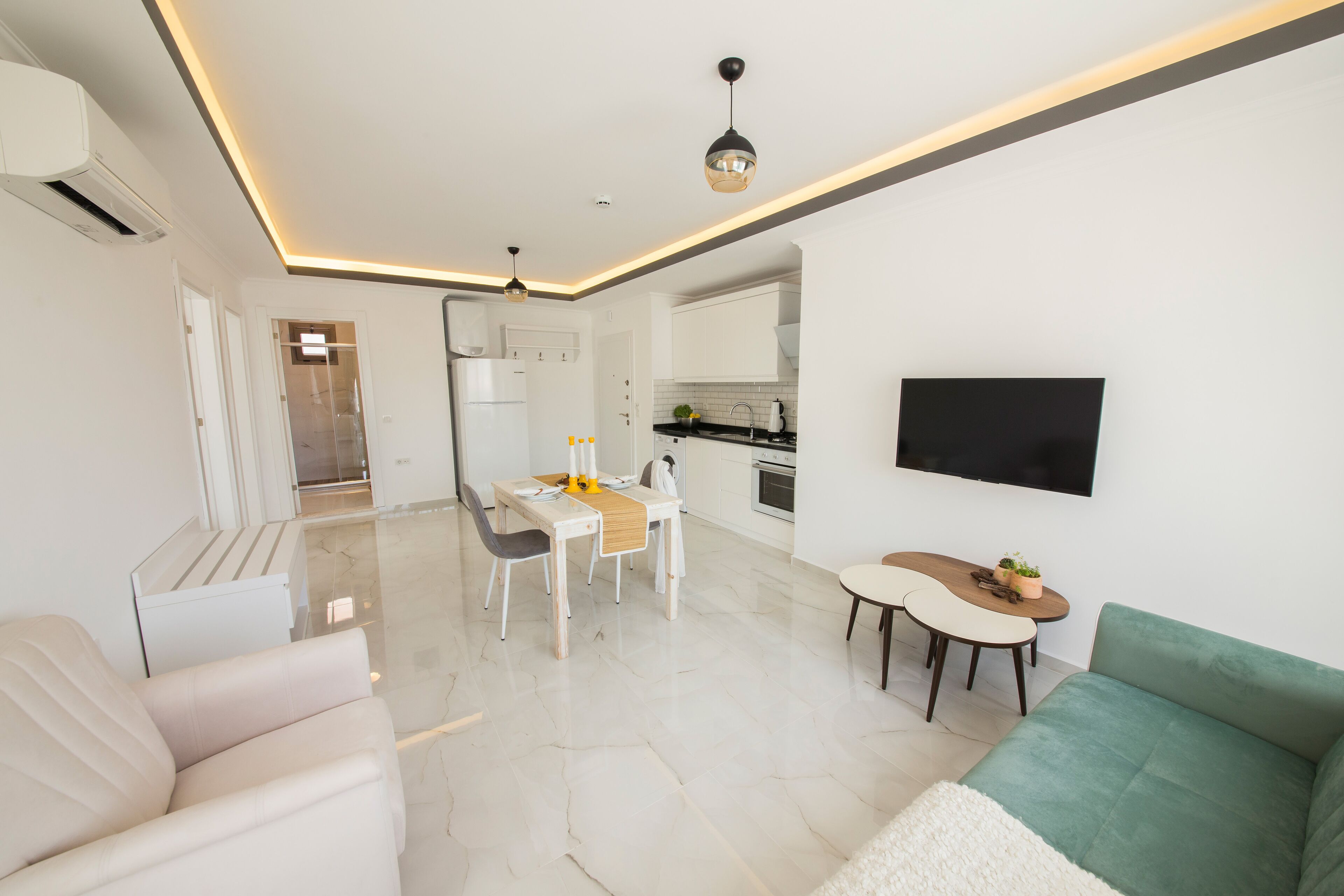 Uma sala de estar moderna com um sofá, uma área de jantar com mesa e cadeiras, uma cozinha com TV fixada na parede e um corredor que dá para outro quarto.