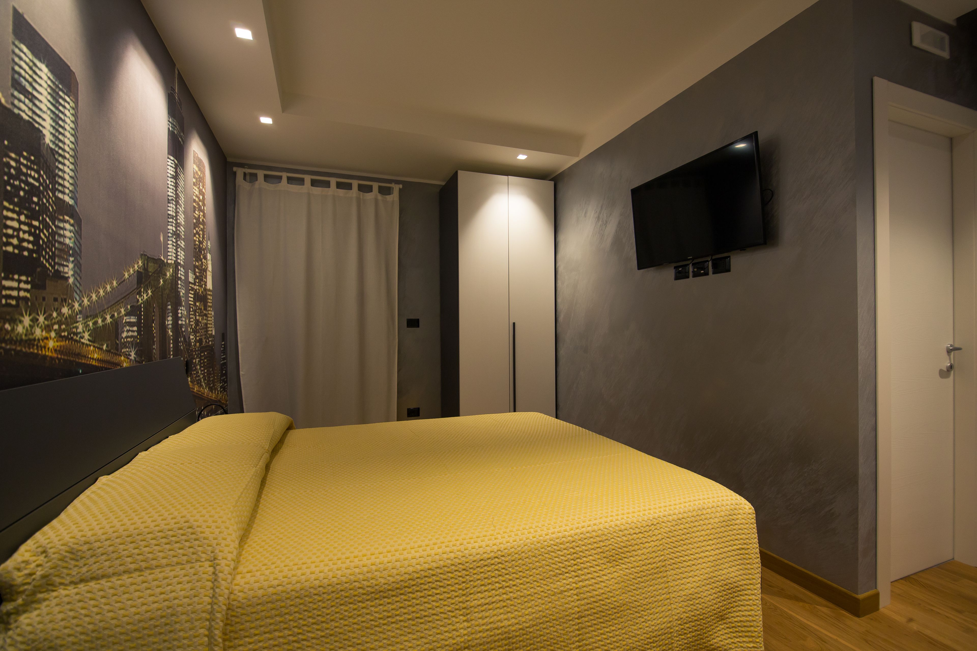 Quarto com cama, TV montada na parede e um mural com vista da cidade.