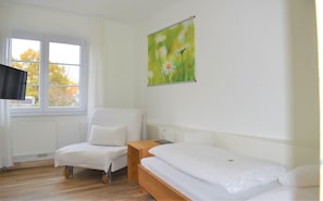 Quarto de hotel com uma cama de solteiro, uma cadeira, uma mesinha, televisão e janela com cortinas.