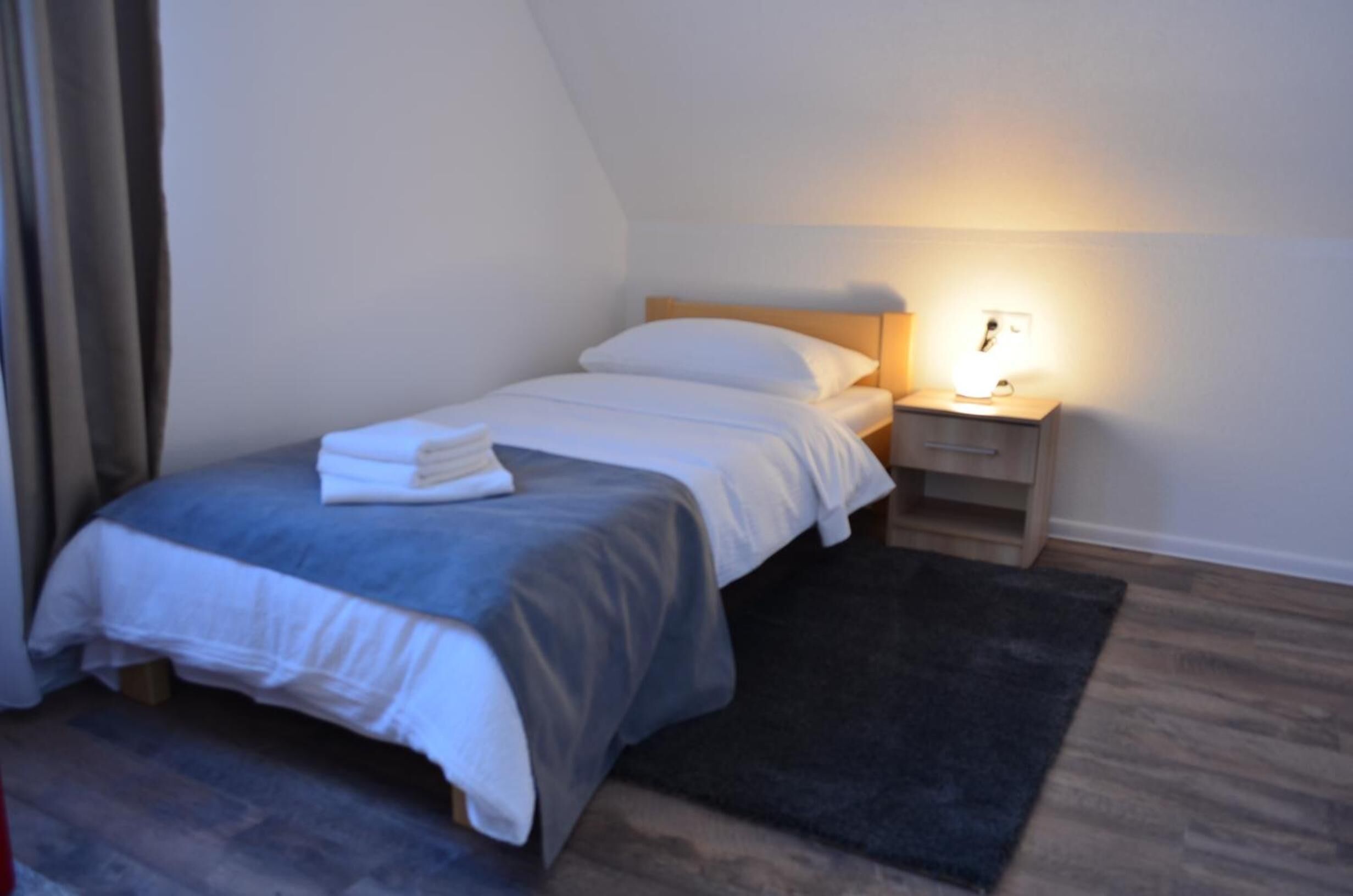 Cama bem arrumada com colcha azul e lençóis brancos, mesa de cabeceira com abajur e tapete escuro sobre piso de madeira.