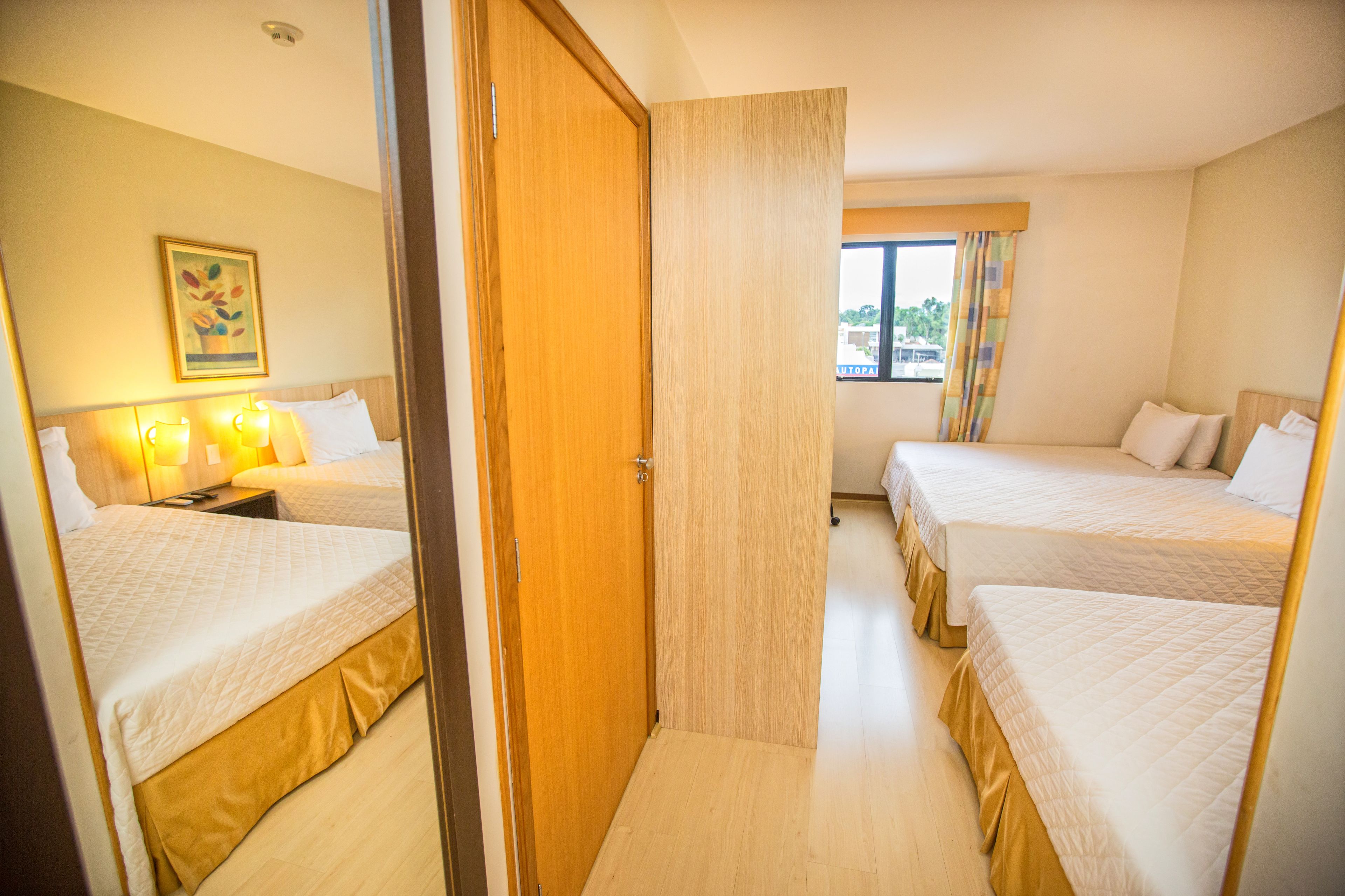 Quarto de hotel com duas camas, uma porta de madeira, uma janela com cortinas e um quadro na parede.