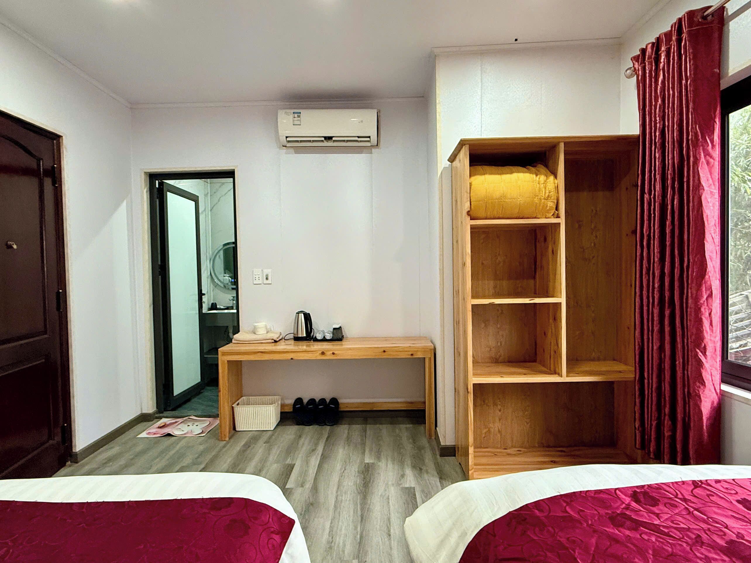 Quarto com duas camas, uma escrivaninha de madeira, uma prateleira, ar condicionado e uma janela com cortinas.