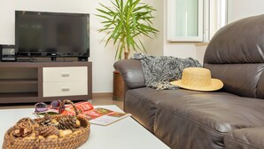 Uma sala de estar com um sofá de couro marrom, uma televisão em um móvel, um vaso de planta e uma cesta de vime com objetos sobre uma mesa branca.